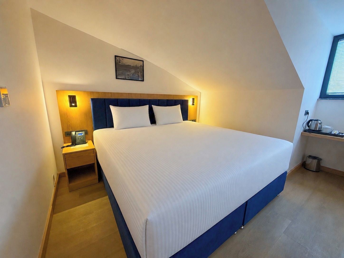 Quarto de hotel com cama grande, cabeceira de madeira, abajures, uma mesinha com chaleira e um quadro na parede.