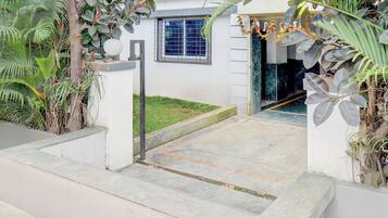 Entrada de casa com caminho de concreto que leva a porta, cercada por vegetação exuberante e um pequeno gramado.