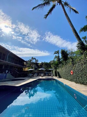 Piscina com piso ladrilhado, rodeada por espreguiçadeiras e guarda-sóis, com um edifício com varanda ao fundo.