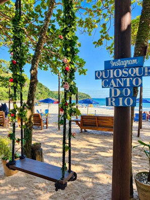 Cena de praia com um balanço, um banco de madeira e uma placa com os dizeres 'Instagram' e 'QUIOSQUE CANTO DO RIO'.