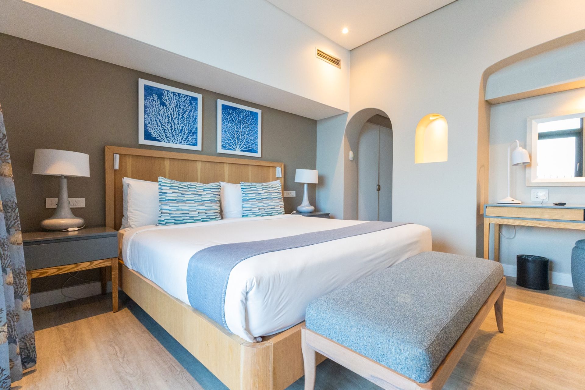 Quarto de hotel com uma cama grande, duas mesas de cabeceira, um banco, um espelho e duas luzes fixadas na parede.