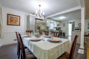 Uma sala de jantar com mesa posta para quatro pessoas, um candelabro e uma cozinha ao fundo.
