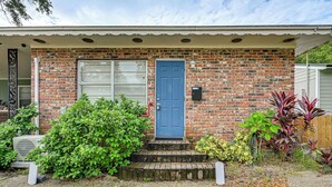 Uma casa de dois andares em tijolo com porta da frente azul, um pequeno alpendre e um jardim com diversas plantas.