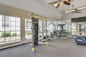Academia com diversos equipamentos de ginástica, janelas amplas e um ventilador de teto.