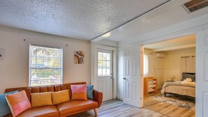 Quarto com um sofá de couro marrom, piso de madeira e porta que dá para um quarto com cama e mesa de cabeceira.