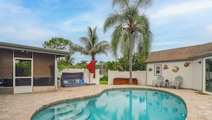 Piscina com borda azulejada, área de estar coberta com sofá e almofadas, e um pequeno deck com cadeiras.