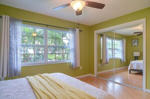 Quarto com cama, ventilador de teto, uma janela grande com cortinas e um espelho refletindo o ambiente.