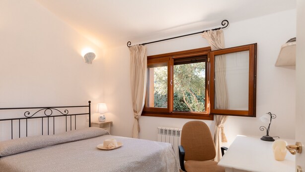 Quarto com cama, escrivaninha, cadeira, janela com cortinas, radiador e luminária na parede.