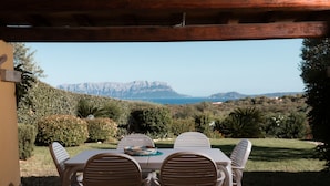 Um pátio externo com mesa e cadeiras, com vista para uma paisagem montanhosa e o mar.