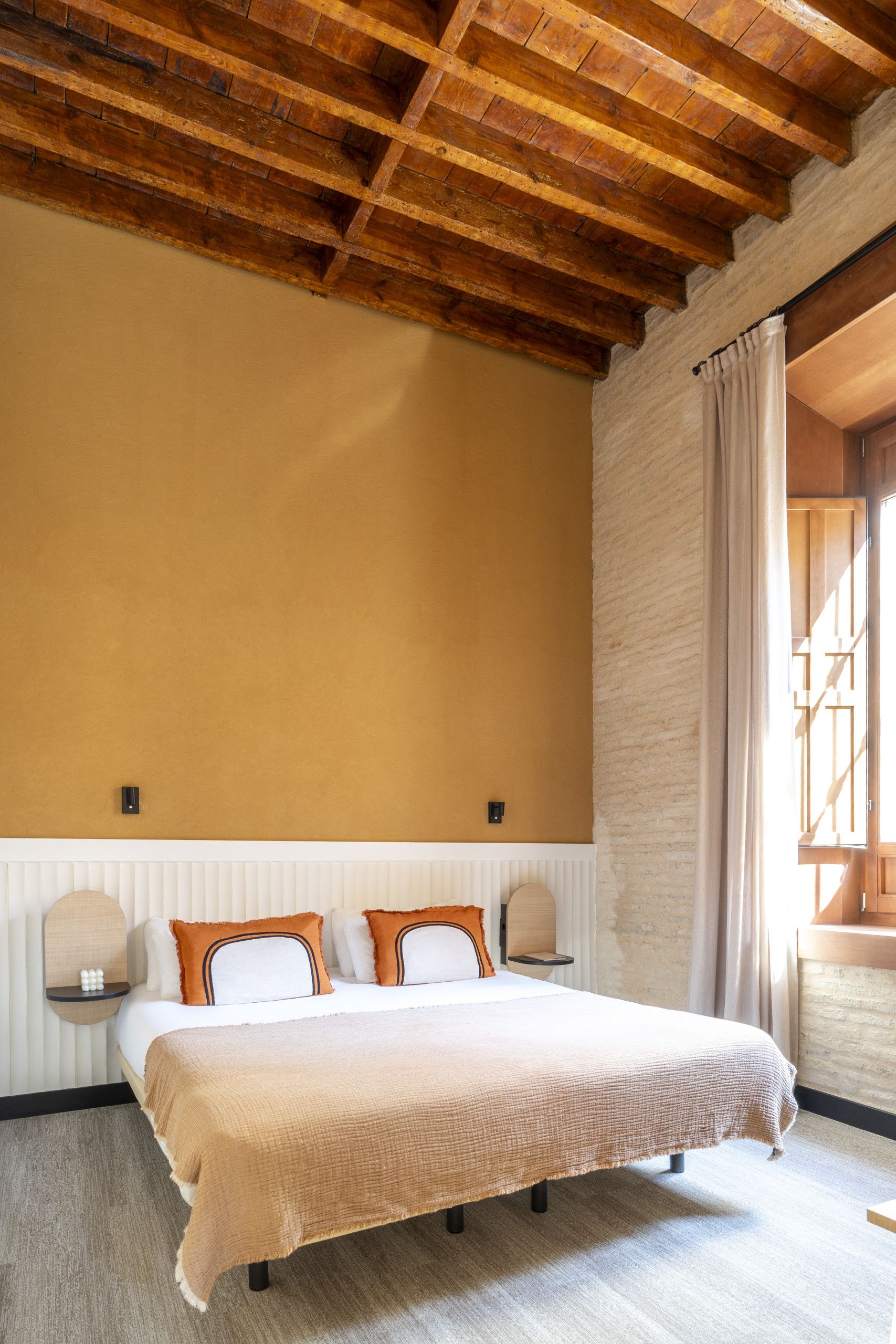 Quarto com teto de madeira, cama com roupa de cama branca e laranja, mesa de cabeceira e janela com cortinas.