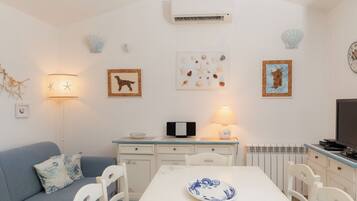 Quarto com mesa branca, cadeiras, sofá azul, televisão e objetos decorativos na parede.