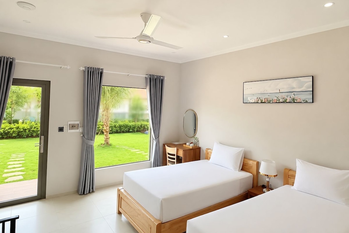 Quarto de hotel com cama, mesas de cabeceira, escrivaninha, cadeira, ventilador de teto e uma grande janela com cortinas.