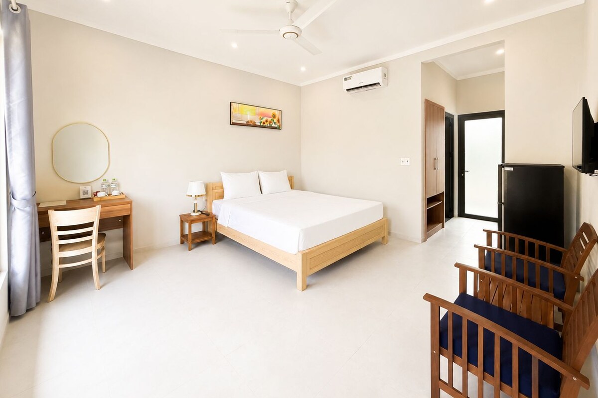 Quarto de hotel com cama, escrivaninha, cadeira, mesa de cabeceira e televisão.