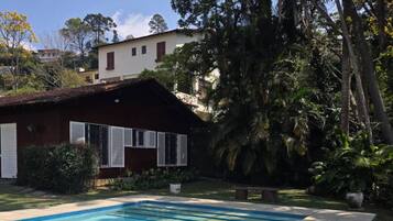 Piscina com deck ladrilhado, rodeada por vegetação exuberante e um edifício com venezianas brancas.