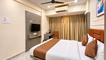 Quarto de hotel com cama, televisão, ar condicionado, escrivaninha com cadeira e janela com cortinas.