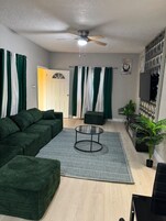 Sala de estar com sofá seccional, mesa de centro de vidro, ventilador de teto e tapete estampado.
