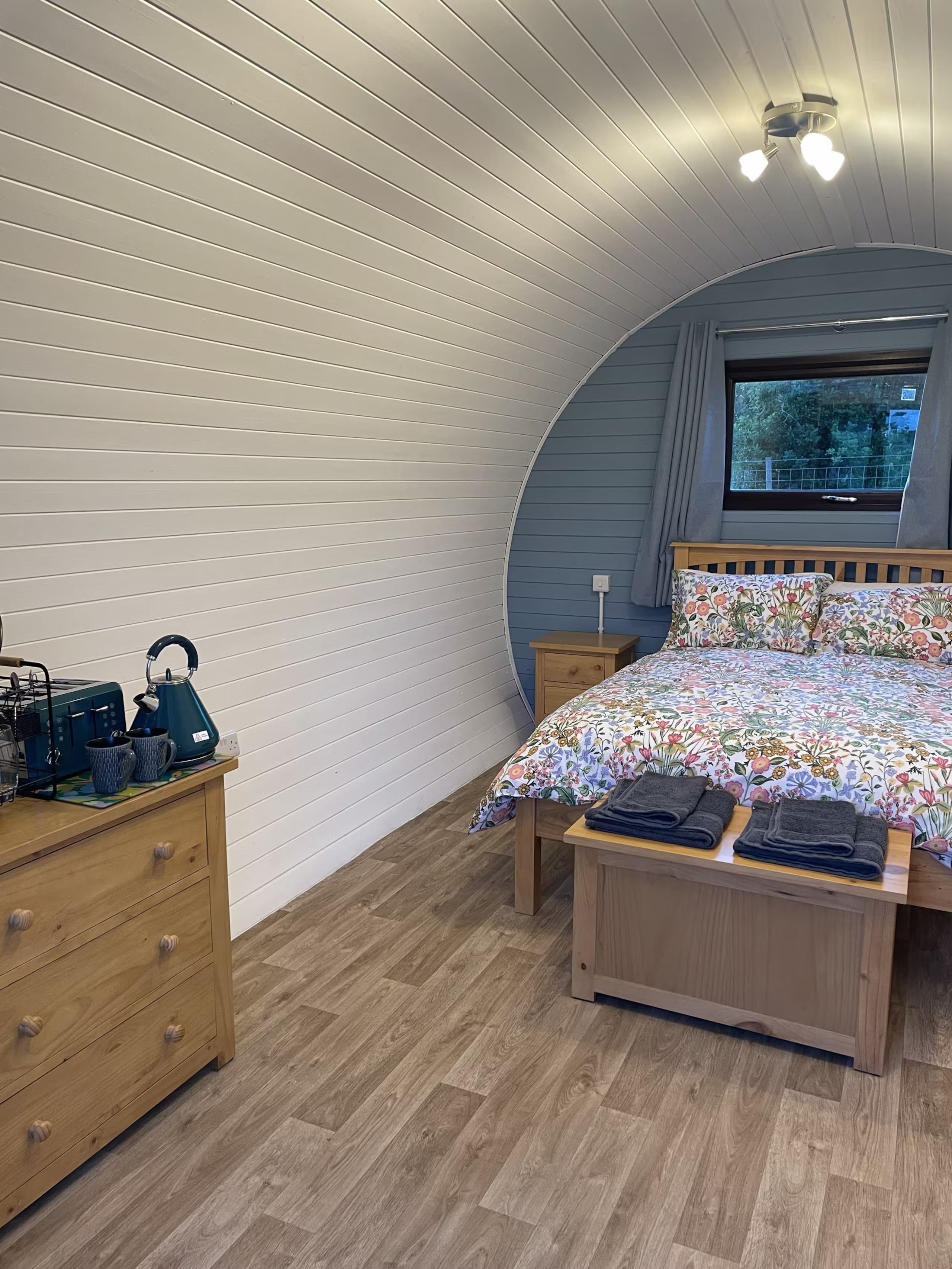 Quarto compacto com cama, cômoda de madeira, mesinha com toalhas e janela com vista para área verde.