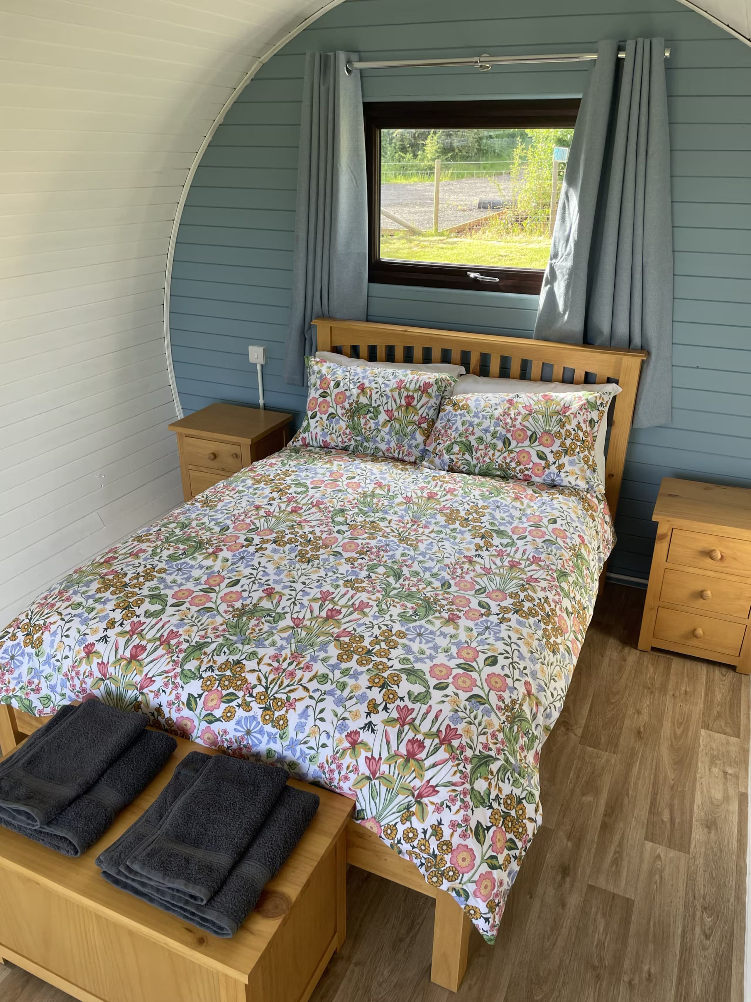 Quarto com cama de madeira, roupa de cama floral, mesa de cabeceira de madeira e janela com cortinas.