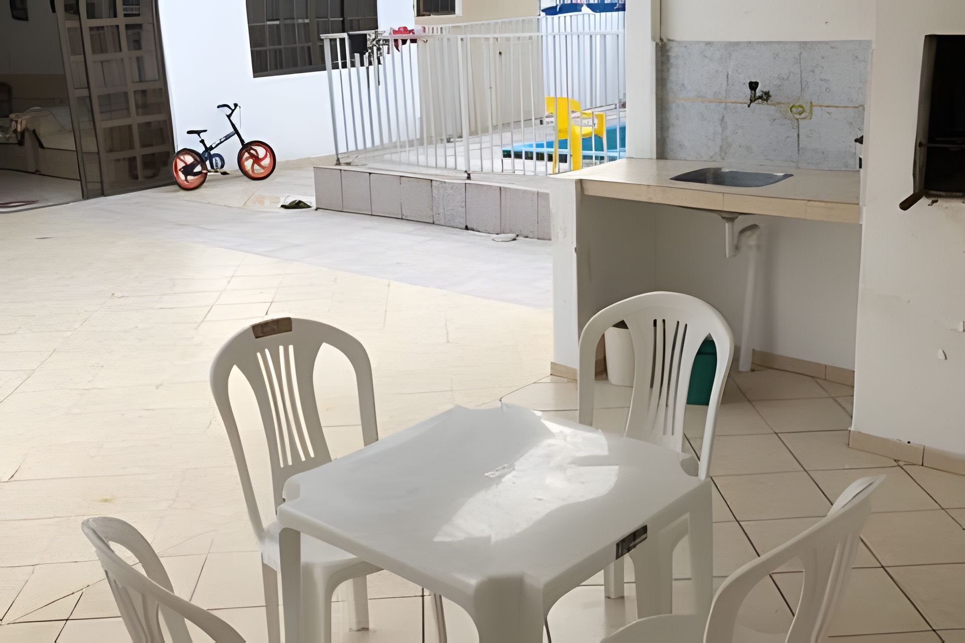 Quarto com uma mesa e cadeiras brancas, pequena área de cozinha com pia e vista para uma varanda com uma bicicleta.