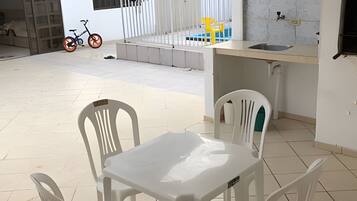 Quarto com uma mesa e cadeiras brancas, pequena área de cozinha com pia e vista para uma varanda com uma bicicleta.