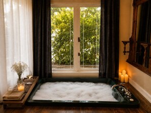 Um quarto de spa com uma grande banheira cheia de bolhas, uma janela com cortinas e velas ao lado.