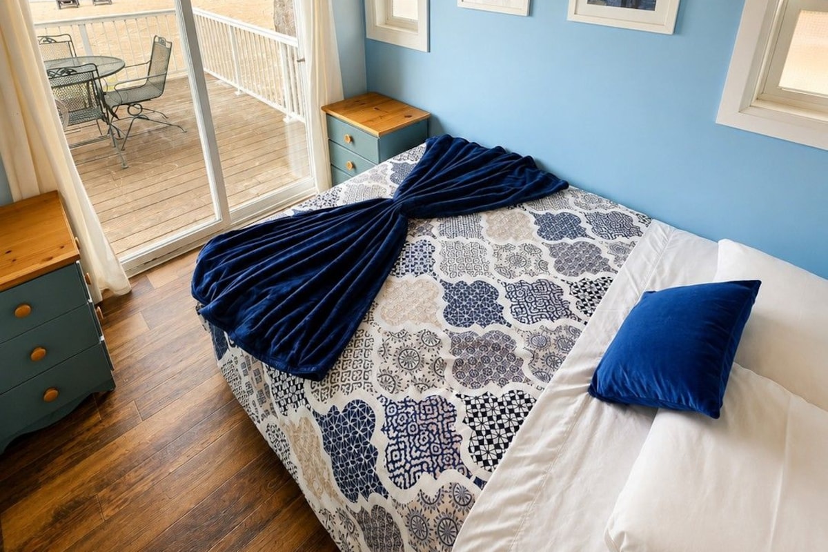 Quarto com cama, mesa de cabeceira, janela e vista para área externa com piscina e cadeiras.
