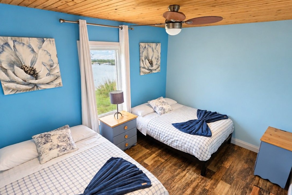 Quarto com duas camas, piso de madeira, ventilador de teto e quadros nas paredes.