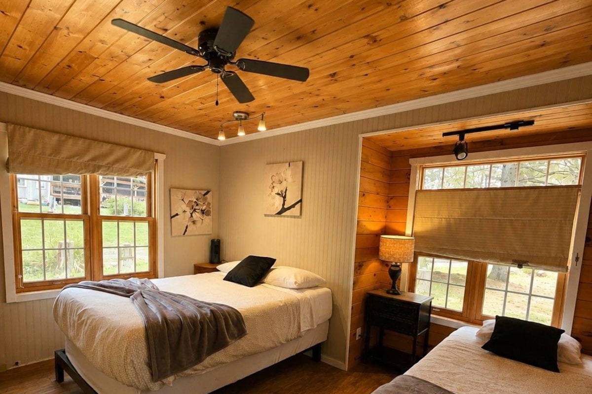 Quarto com duas camas, piso de madeira, ventilador de teto e janelas com persianas.