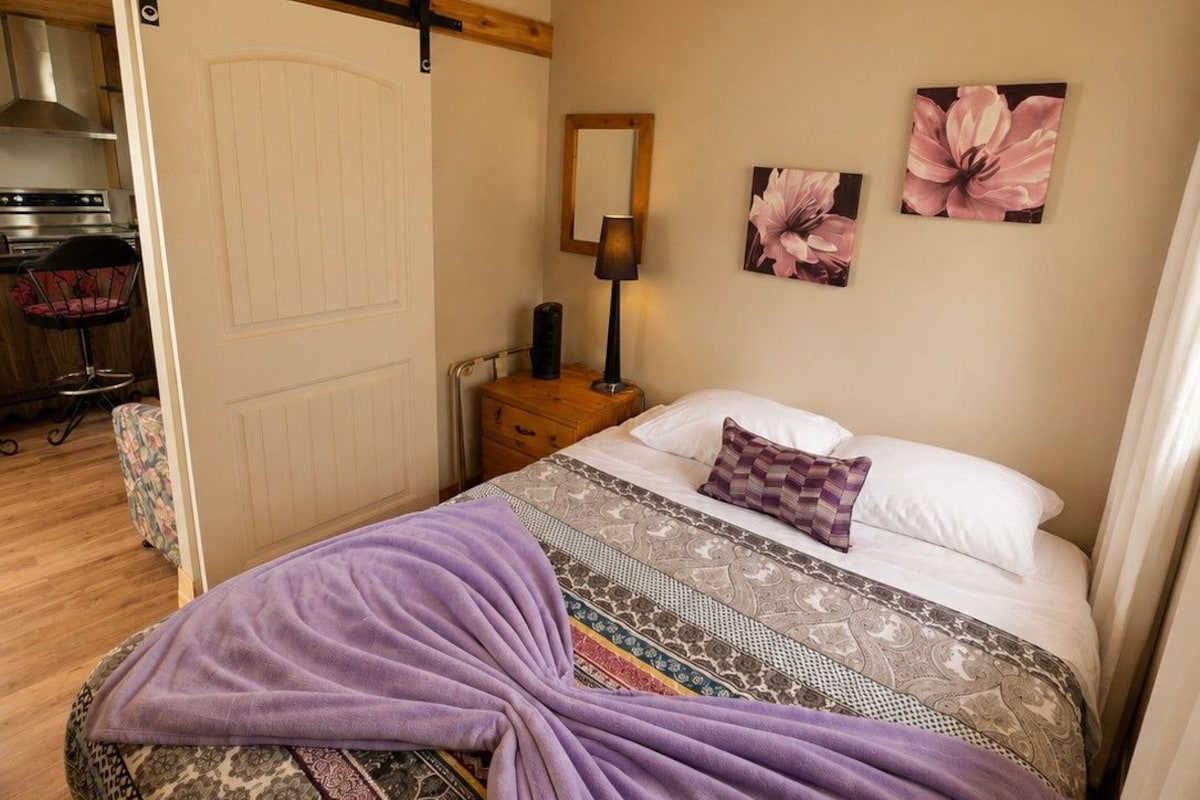 Quarto com teto de madeira, cama com colcha estampada, manta roxa, mesa de cabeceira com abajur e parede com dois quadros florais.