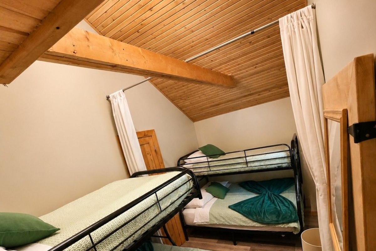 Quarto pequeno com duas beliches, vigas de madeira no teto, uma mesa de madeira e uma janela com cortinas.
