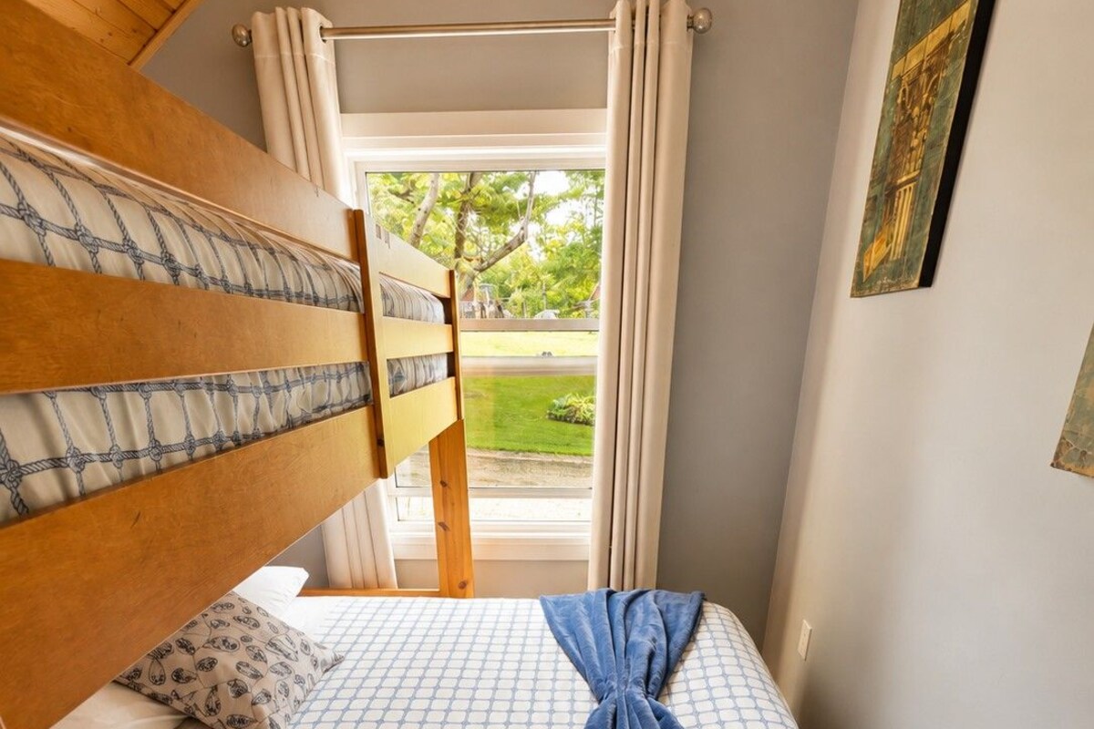 Um quarto com uma cama beliche, uma janela com cortinas, uma cama com colcha estampada e piso de madeira.