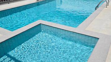 Piscina retangular com borda azulejada, rodeada por uma cerca de metal, e uma pequena parte de um edifício com janelas e uma porta ao fundo.