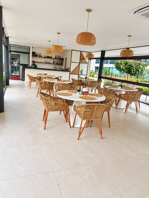 Uma sala de jantar moderna com cadeiras de vime e mesas redondas, um bar com prateleiras cheias de garrafas, e grandes janelas que oferecem vista para o verde.
