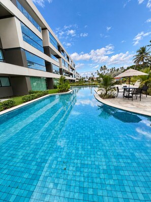 Piscina com piso em azulejo, rodeada por um edifício moderno com vários andares e grandes janelas.