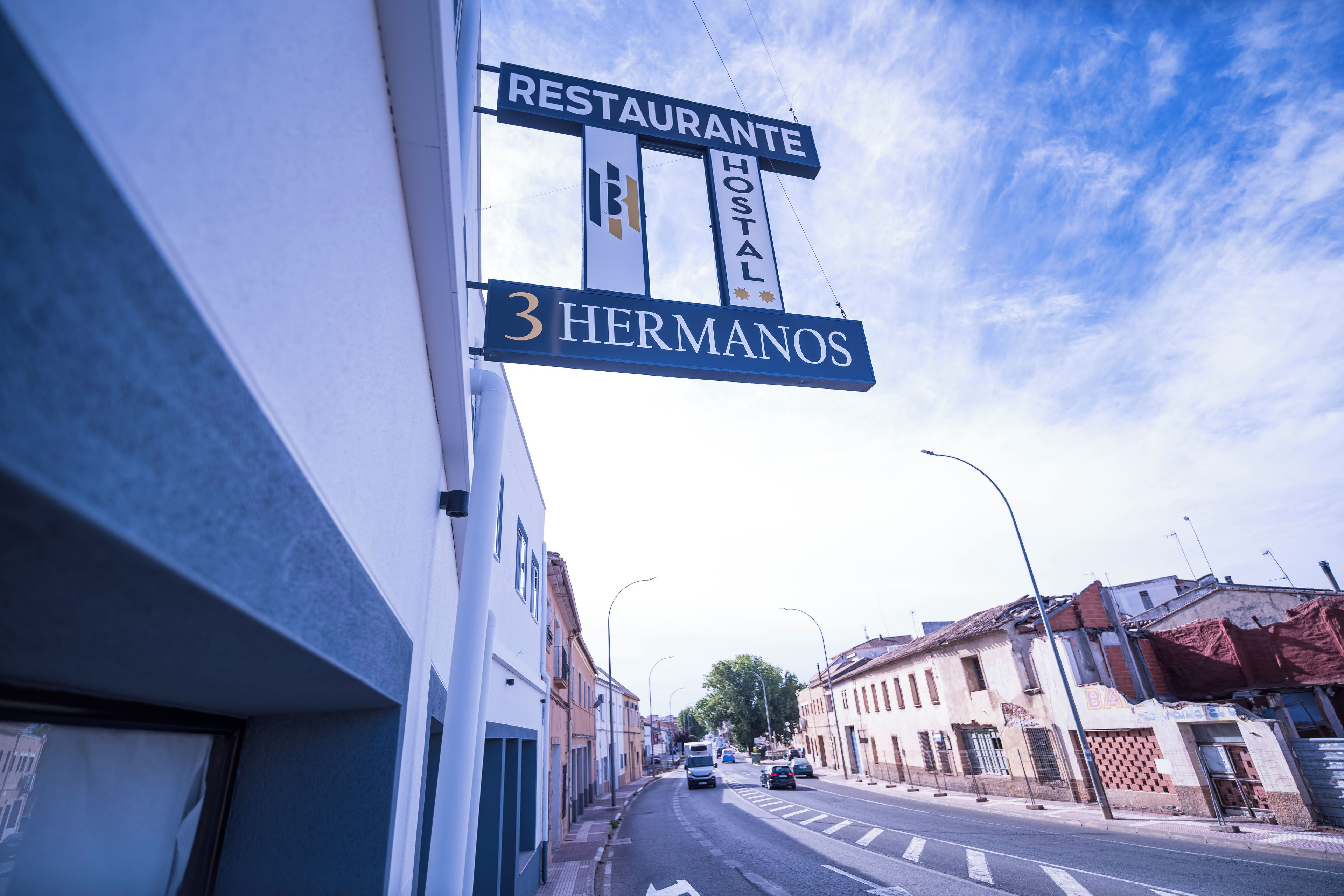 Vista da rua com placa indicando 'RESTAURANTE HOSTAL 3 HERMANOS' e um edifício com entrada visível.
