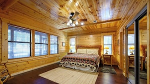 Quarto de cabana de toras com uma cama, ventilador de teto e janelas amplas.