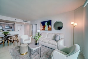 Uma sala de estar moderna com um sofá, cadeiras, uma mesa de centro e um espaço de refeições com mesa e cadeiras. É possível ver uma cozinha ao fundo, com um micro-ondas e armários.