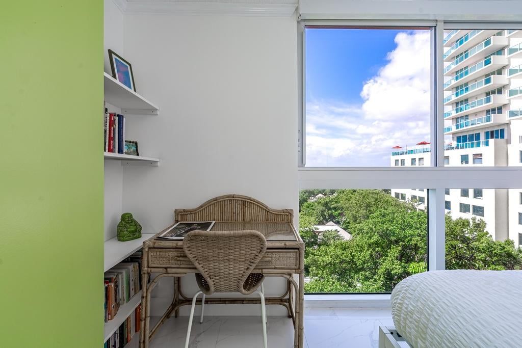 Quarto com escrivaninha, cadeira e vista para um edifício através de uma janela.