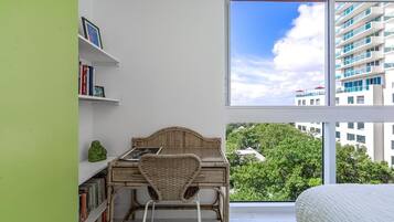 Quarto com escrivaninha, cadeira e vista para um edifício através de uma janela.