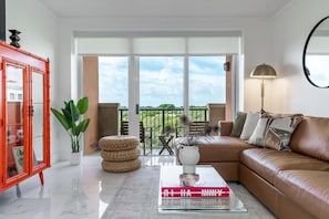 Sala moderna com sofá de couro marrom, mesa de centro de vidro e uma grande janela com vista para o verde.