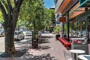 Uma calçada com uma árvore, carros estacionados e uma loja com mesas ao ar livre.