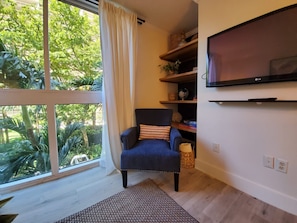 Quarto com uma janela grande, uma poltrona azul, um tapete tecido, uma estante com objetos decorativos e uma TV na parede.