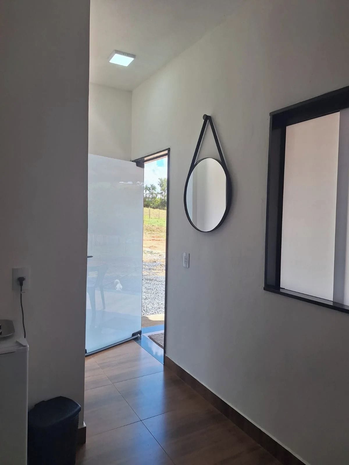 Corredor com porta branca que dá para o exterior, espelho redondo na parede e janela com moldura preta.