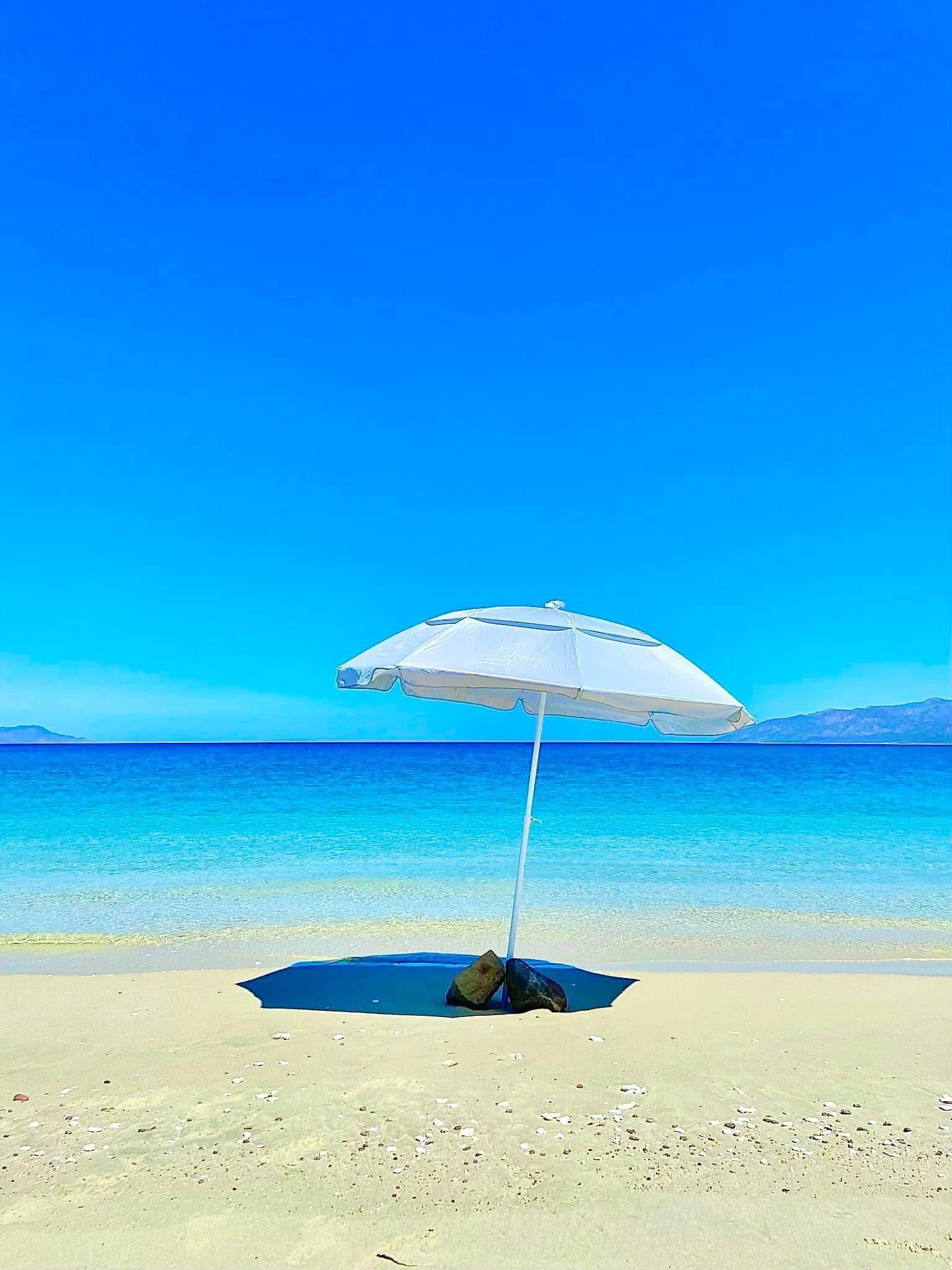 Cena de praia com céu azul límpido, areal dourado e um guarda-sol branco a dar sombra.