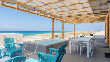 Uma área de estar coberta ao ar livre, com piscina, cadeiras e vista para a praia e o mar.