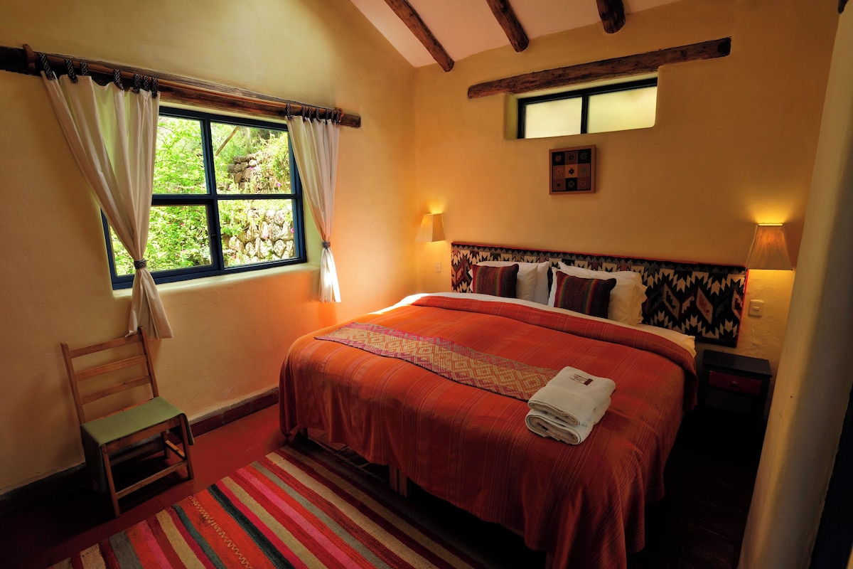 Quarto com cama, janela com cortinas, cabeceira com almofadas estampadas e mesa de cabeceira com abajur.