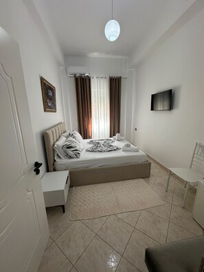 Quarto de hotel com cama, mesa de cabeceira, cadeira, televisão e cortinas.