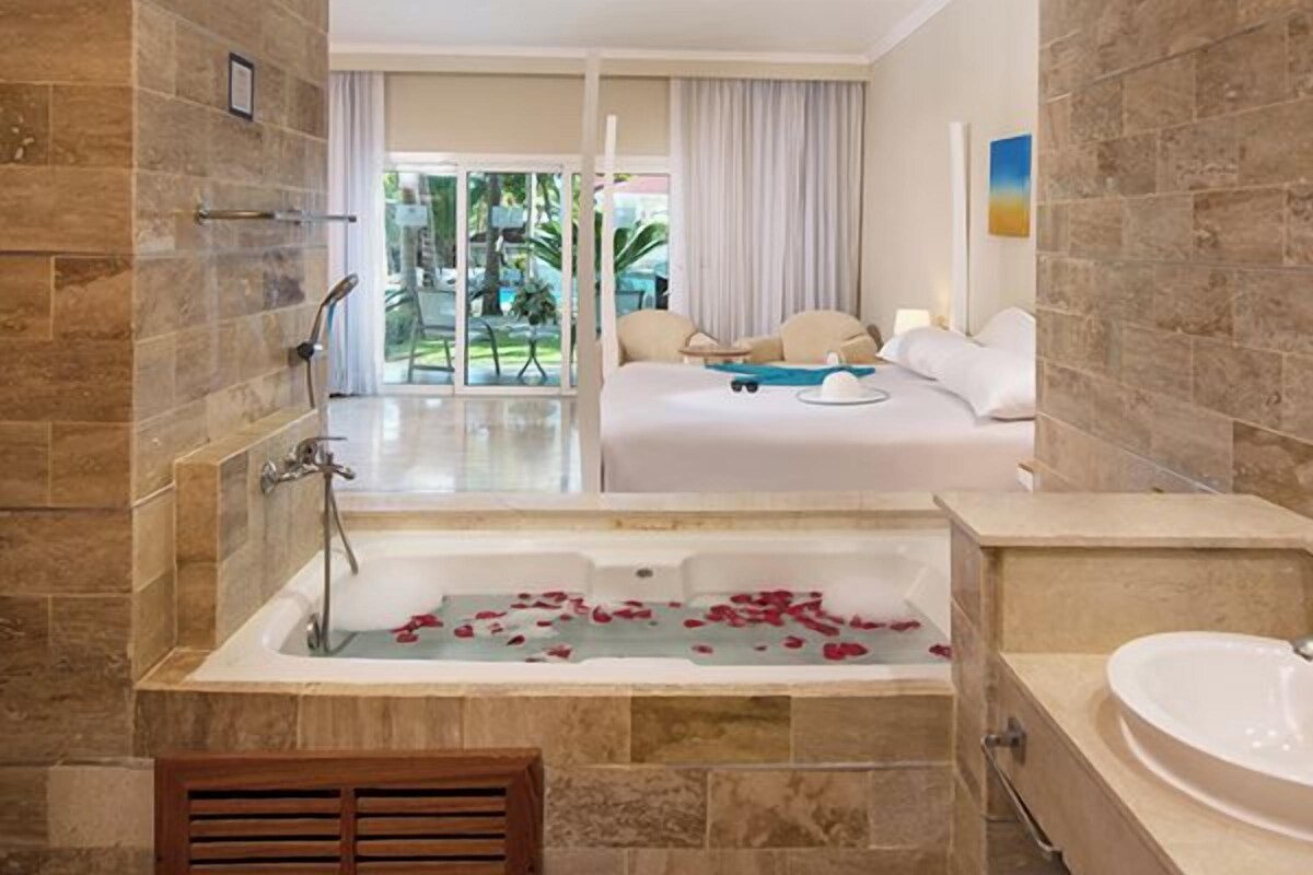 Uma casa de banho moderna com banheira, lavatório e chuveiro. Há uma cama com vista para um jardim ao fundo.