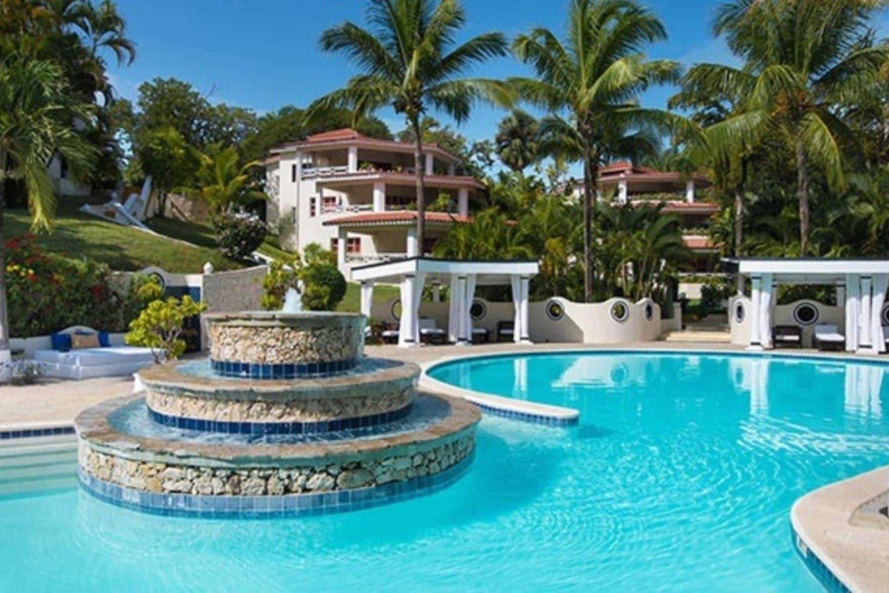 Uma piscina com uma fonte de pedra, cercada por palmeiras e um edifício com varandas.