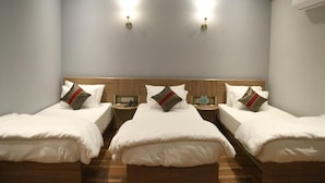 Quarto de hotel com três camas de solteiro, cada uma com cabeceira de madeira, roupa de cama branca e almofadas decorativas. Há mesas de cabeceira com luminárias e um aparelho de ar condicionado fixado na parede.